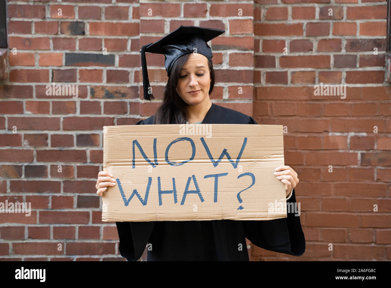 Sad Graduate Student Standing With Now What Placard Against Brick Wall ...