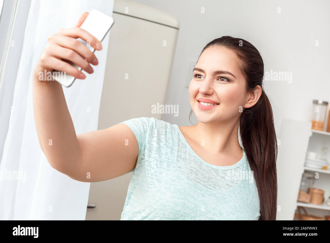 Body Care. Chubby girl standing in kitchen taking selfie on smartphone ...