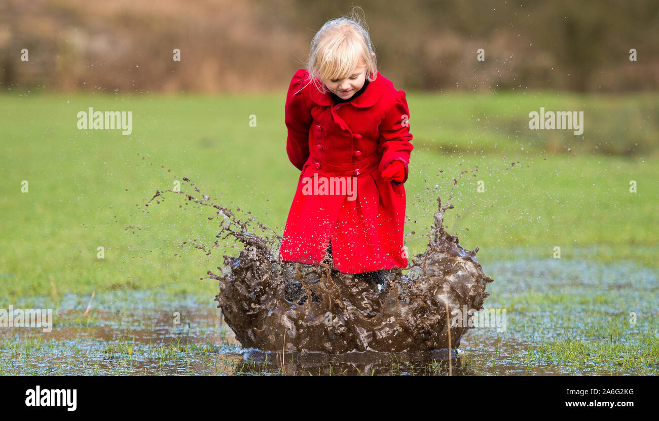 A pretty little blonde girl jumping in a large muddy puddle with a ...