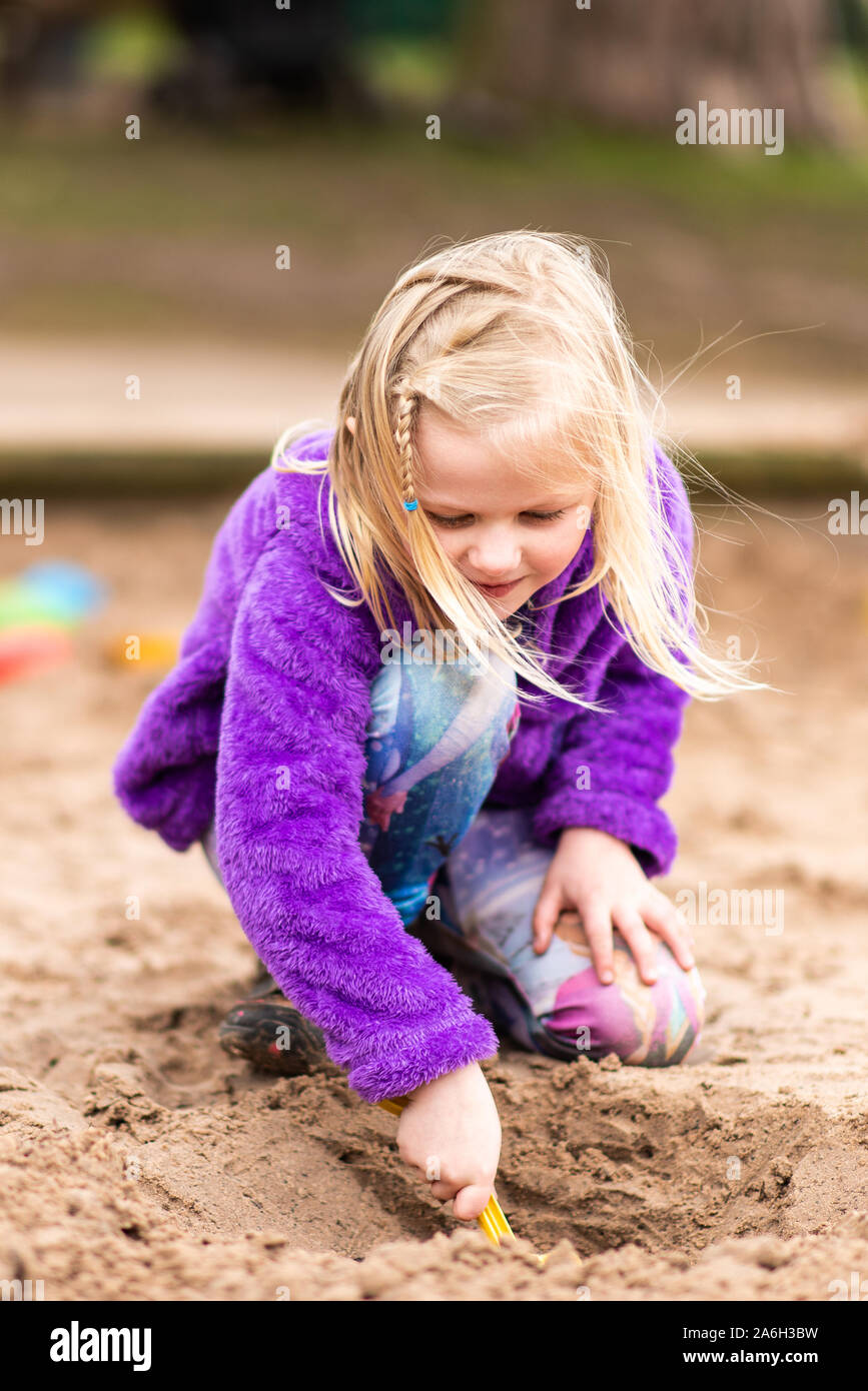 A pretty little girl plays in the sand pit, digging holes and making ...