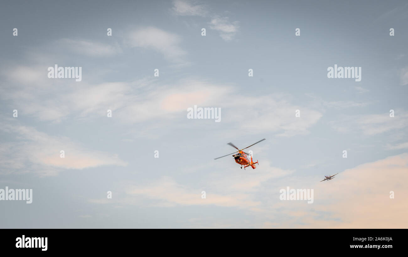 Orange helicopter with airplane behind it flying in a blue sky with ...