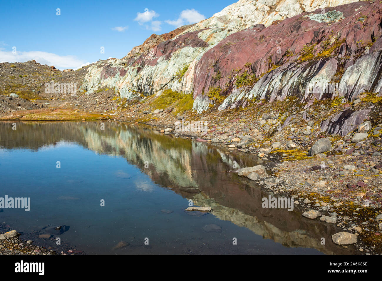 Red, purple and white layered rock cliffs reflected in clear water of a ...