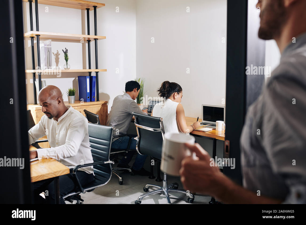 Businessman with mug of coffee entering room with colleagues busy with ...