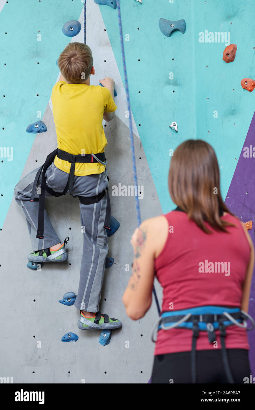 Back view of schoolboy in activewear moving upwards on climbing wall ...