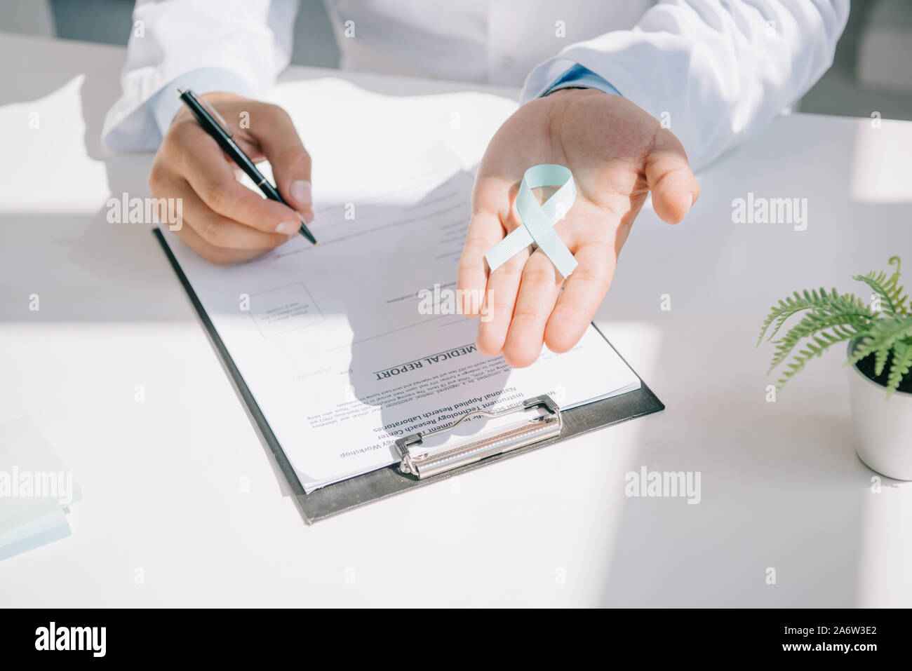 cropped view of doctor holding blue awareness ribbon while writing in ...
