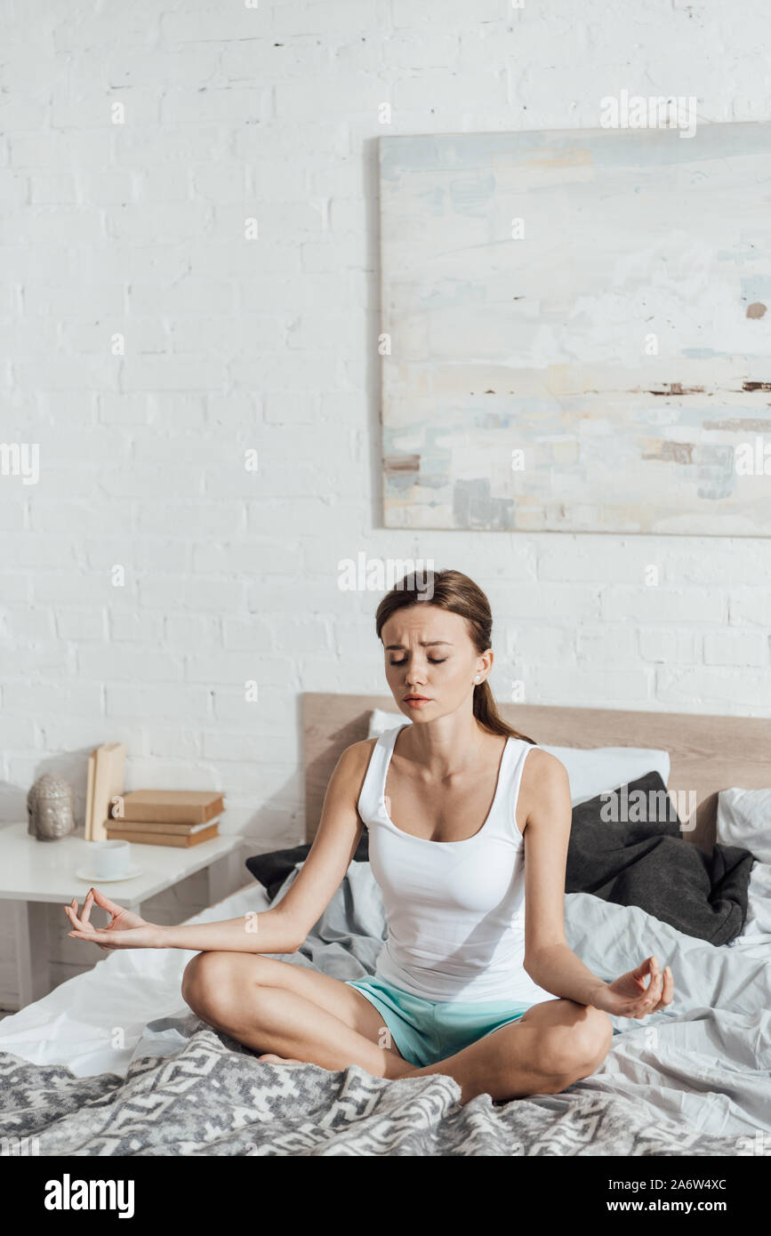 upset young woman sitting on bed in lotus pose with closed eyes Stock ...