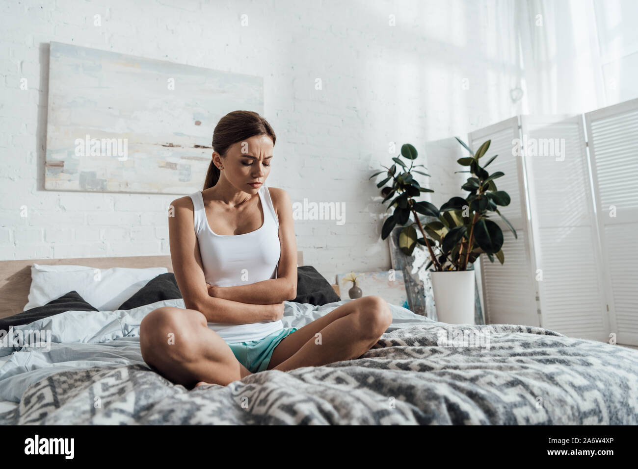 sad young woman sitting on bed and touching belly Stock Photo - Alamy