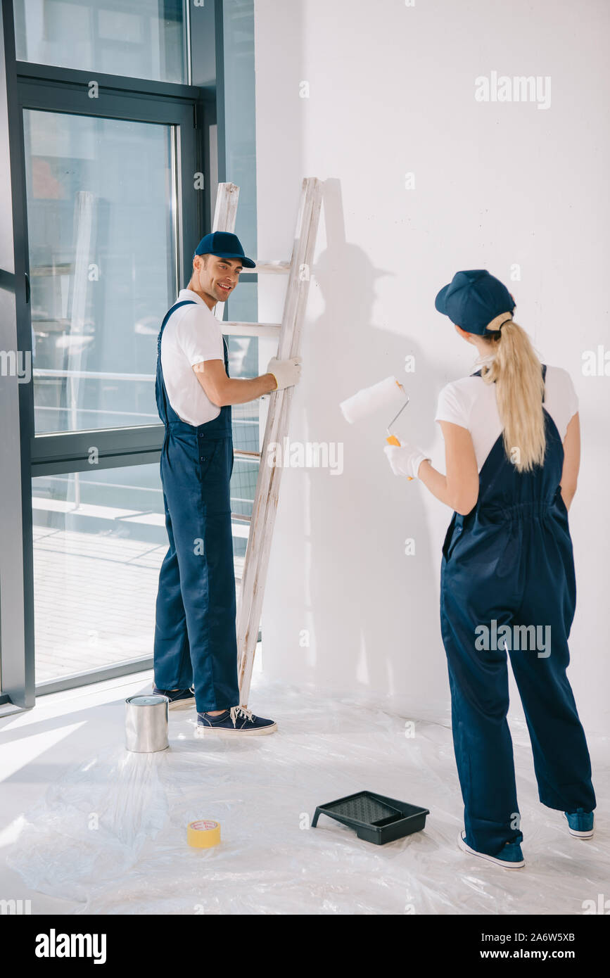 handsome painter standing near ladder while pretty colleague holding ...