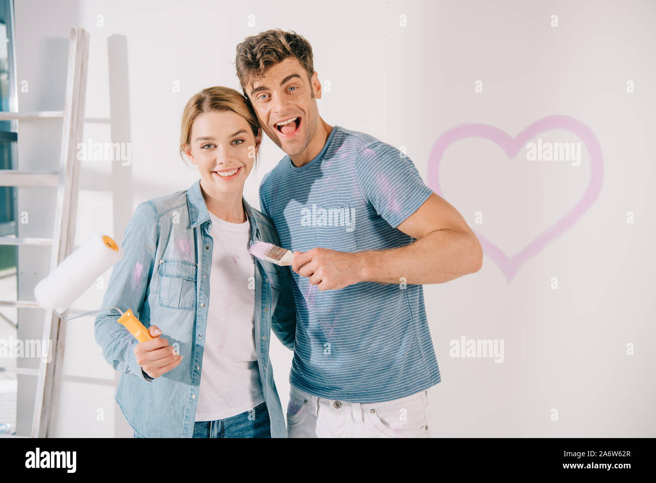 happy young couple looking at camera while holding pink paintbrush and ...