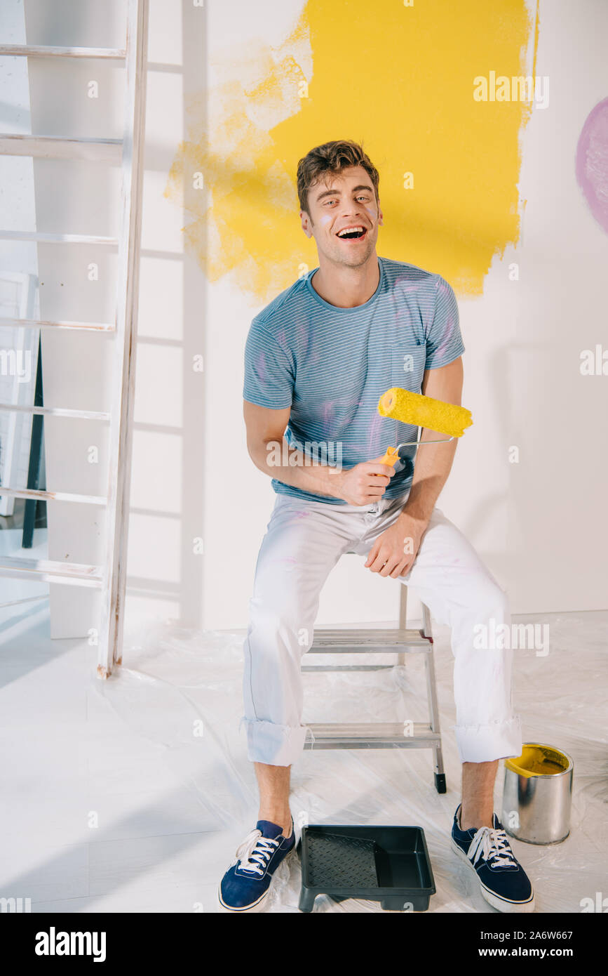 handsome young man sitting on ladder, holding yellow paint roller and ...