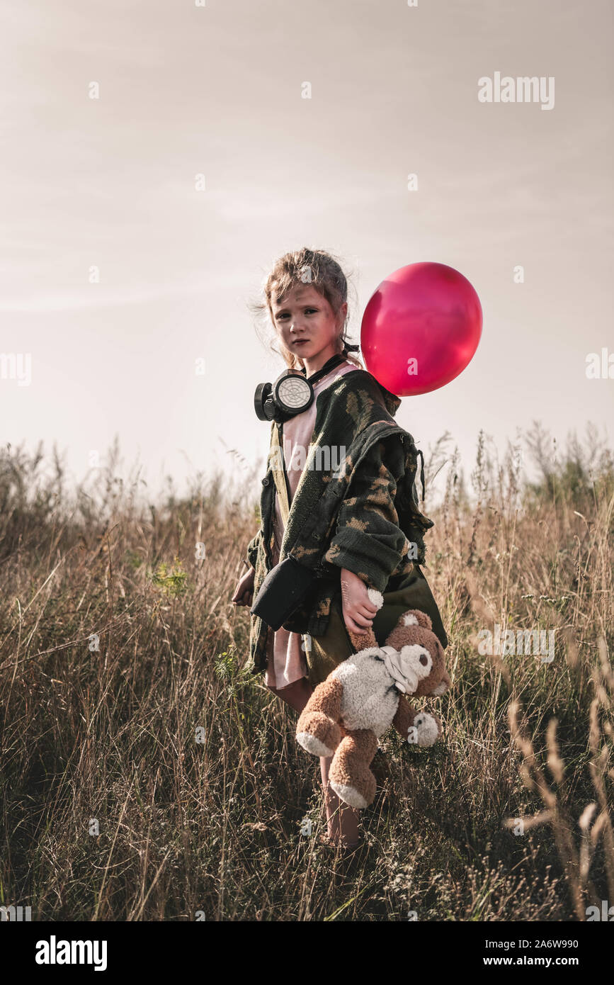 selective focus of cute kid with gas mask and teddy bear holding balloon, post apocalyptic ...