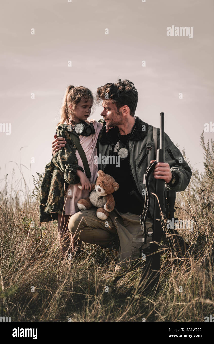 handsome man holding gun near kid with teddy bear in field, post ...