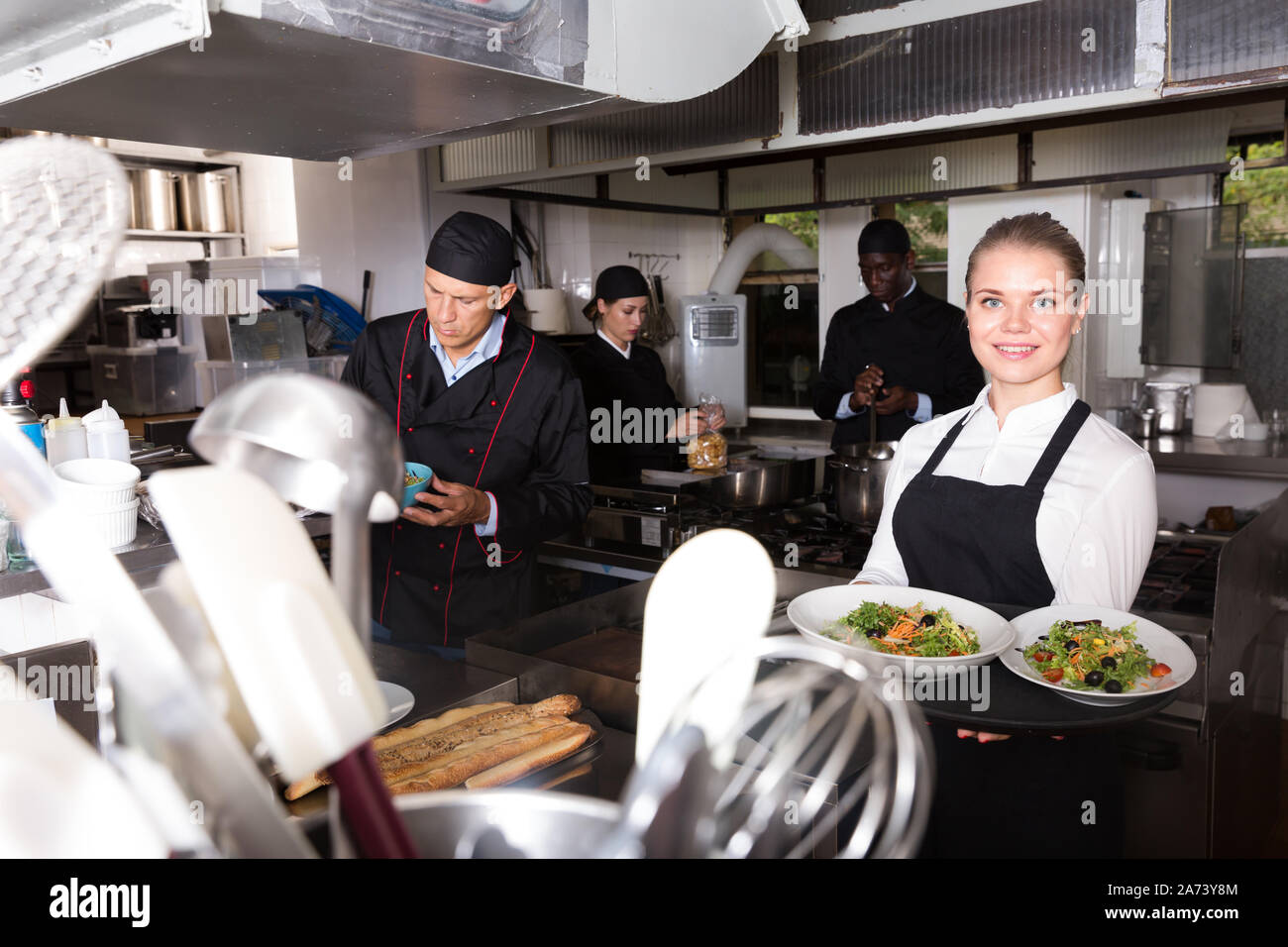 Portrait of attractive waitress with ordered dishes in kitchen of ...