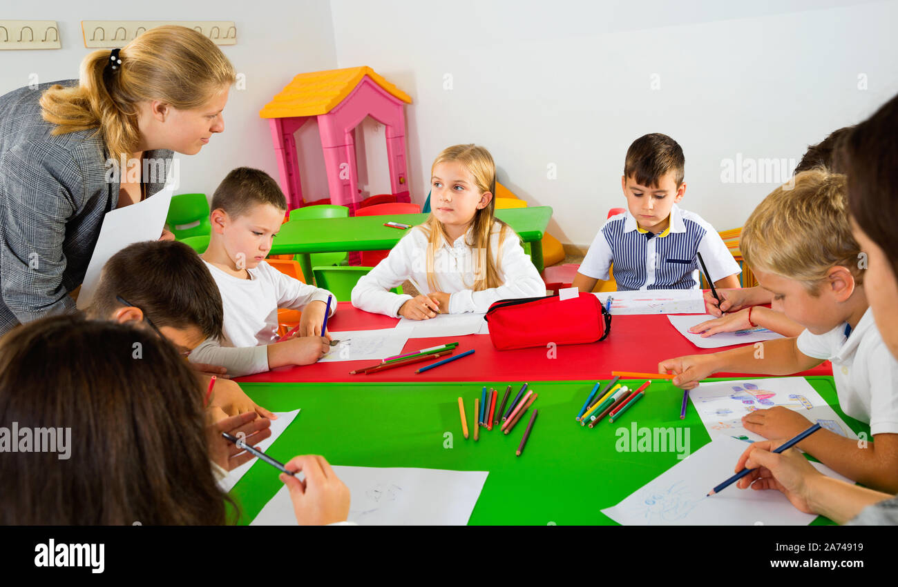 Cheerful female teacher helping schoolkids drawing with color pencils ...