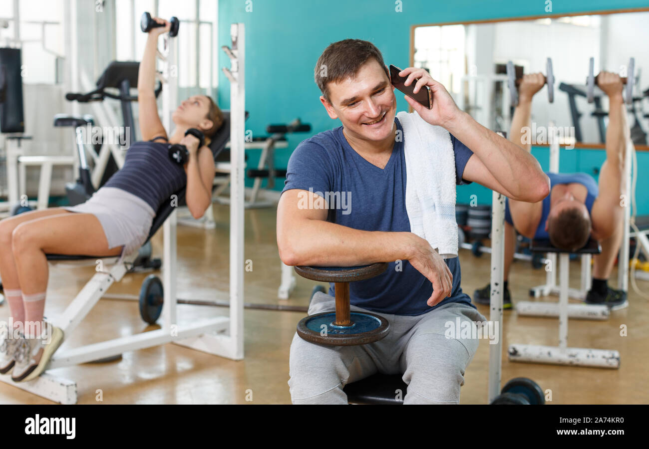 Sporty man sitting on training apparatus and using his gadget in gym ...