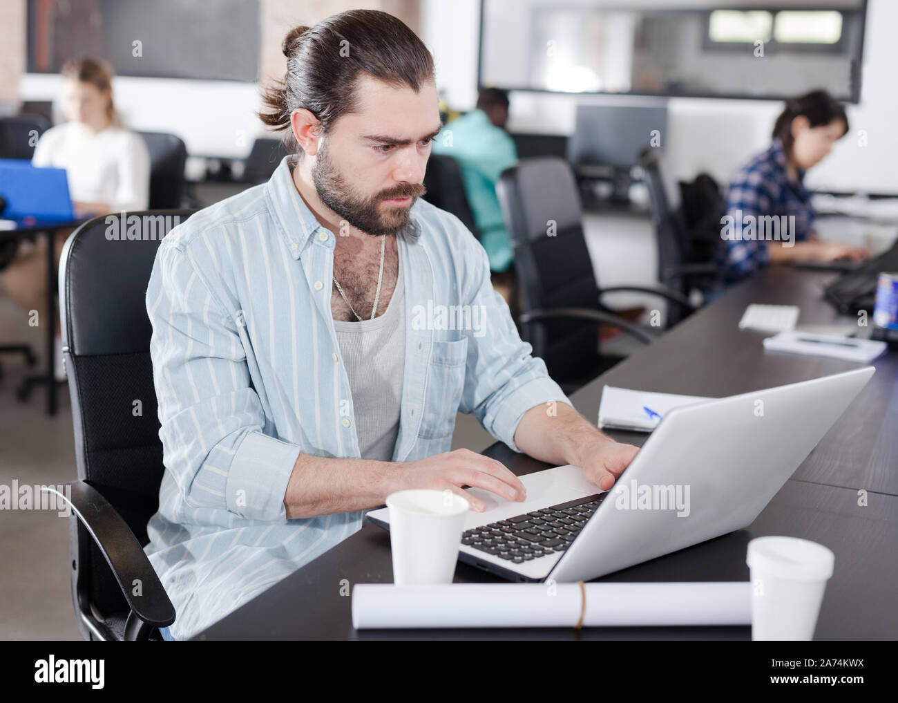 Serious Latin hipster guy working with laptop in busy open plan office ...