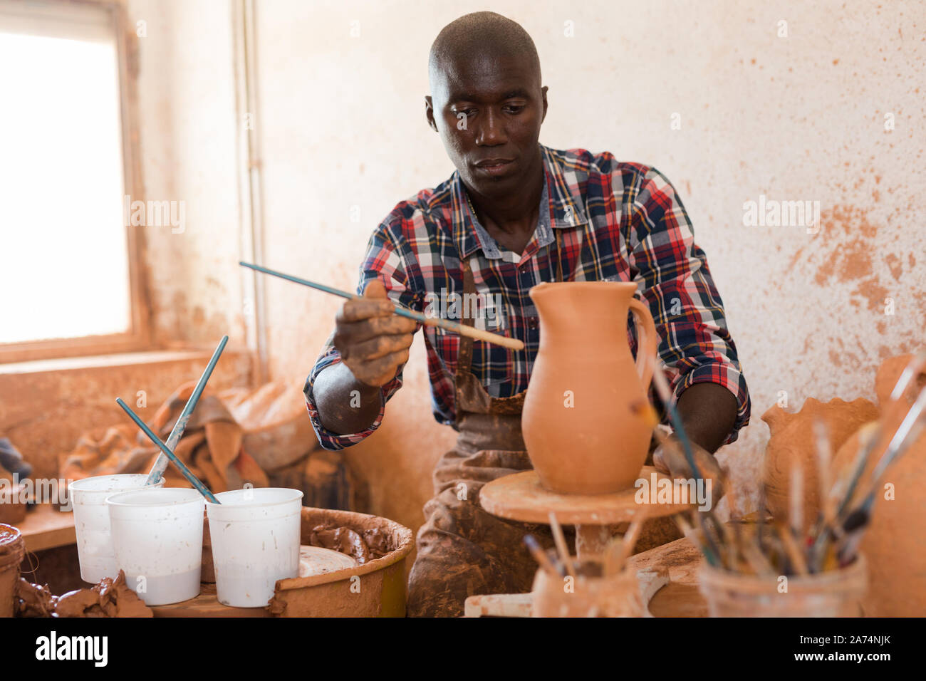Focused African guy working in pottery workshop, painting ceramic jug ...