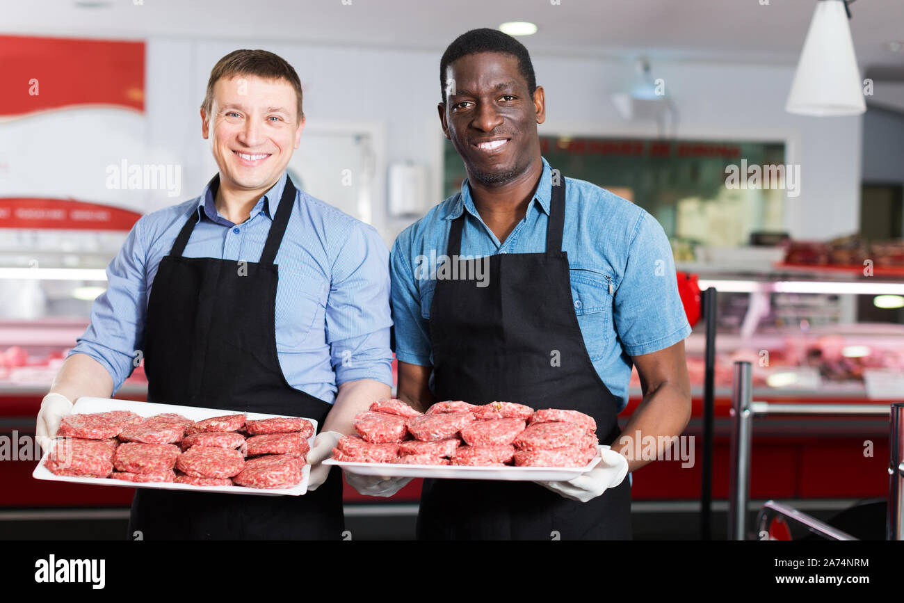 Two happy professional butchers standing in butcher shop with trays of ...