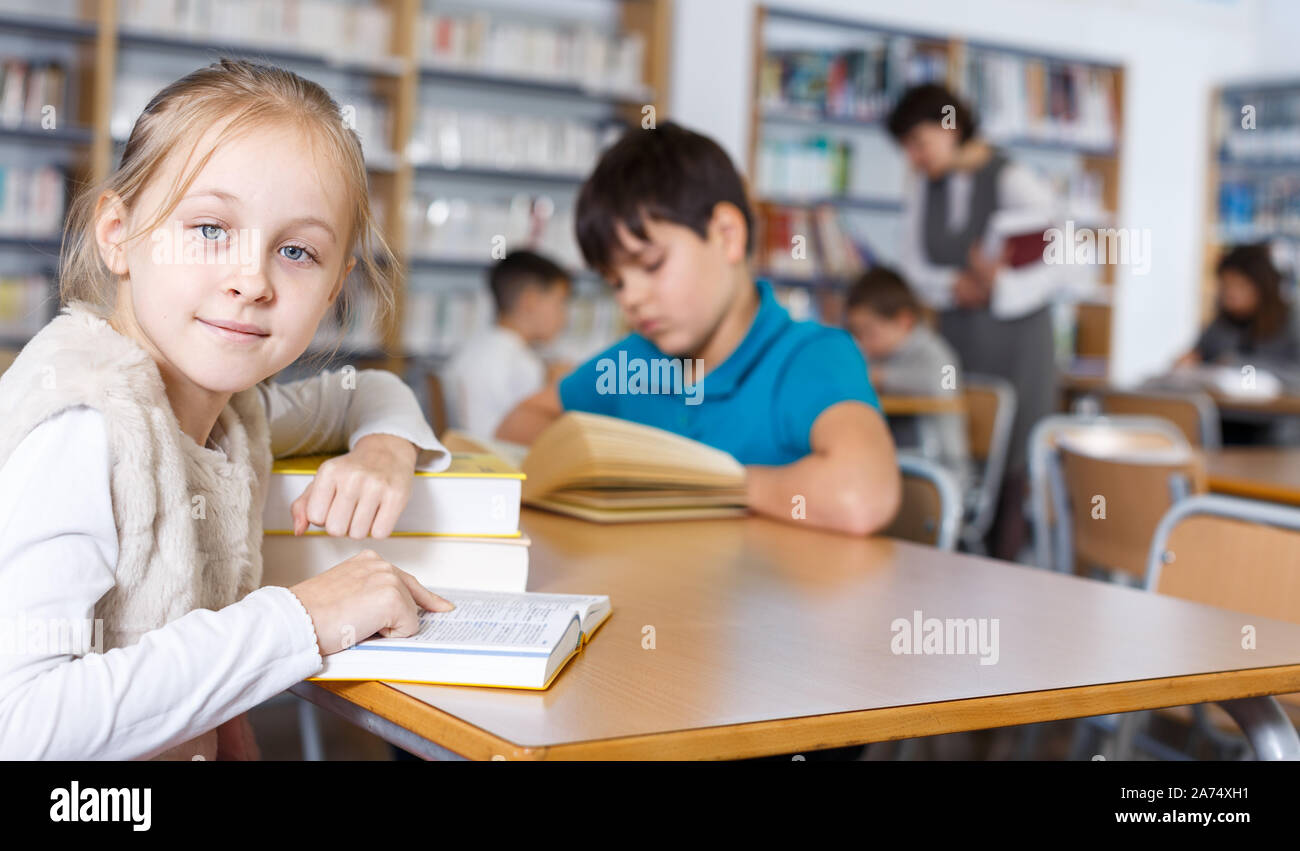 Intelligent tween girl reading in school library on background with ...