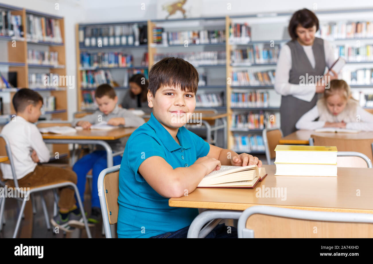 Portrait of smiling intelligent preteen boy reading in school library ...