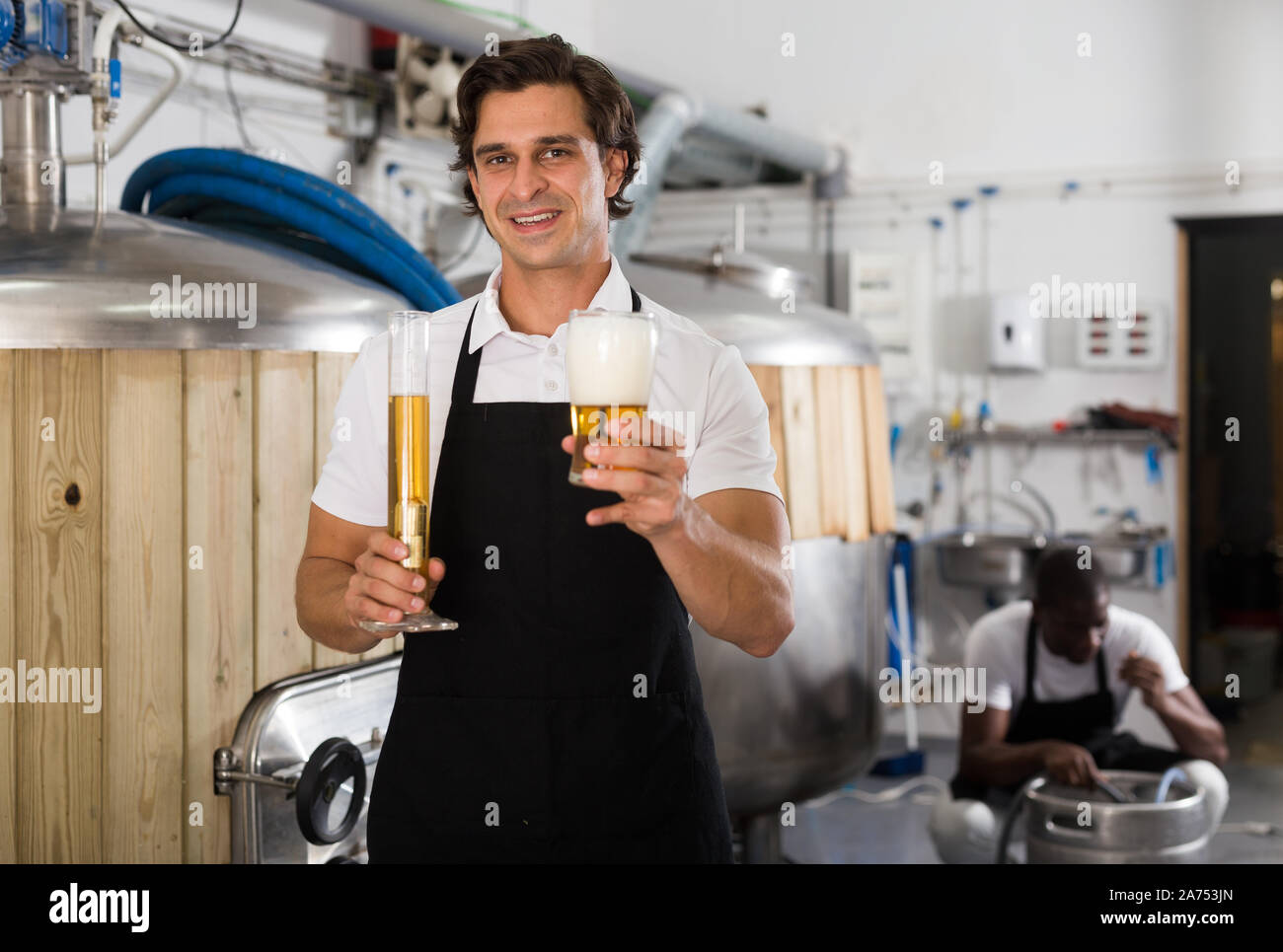 Young man brewer in uniform is standing with glass of beer and flask in ...