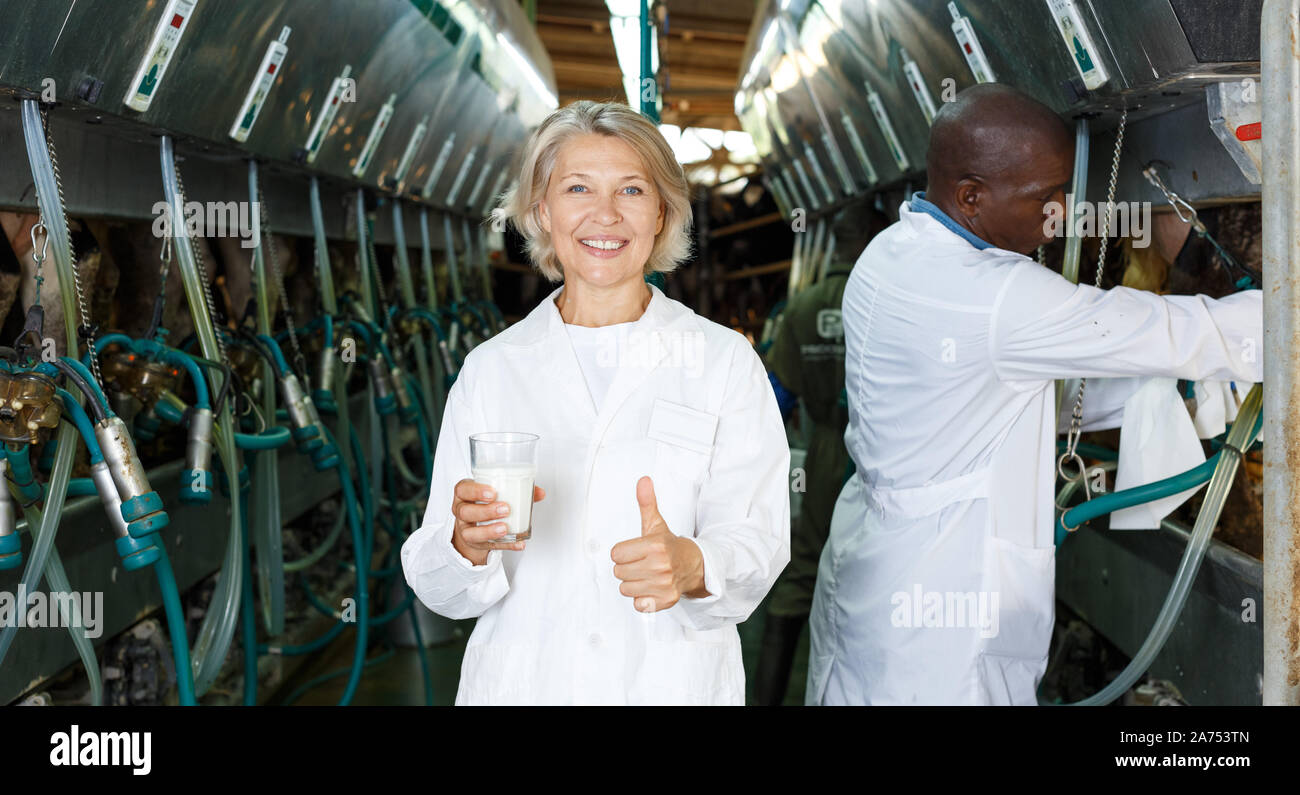 Female dairy engineer in white robe standing with glass of milk near ...
