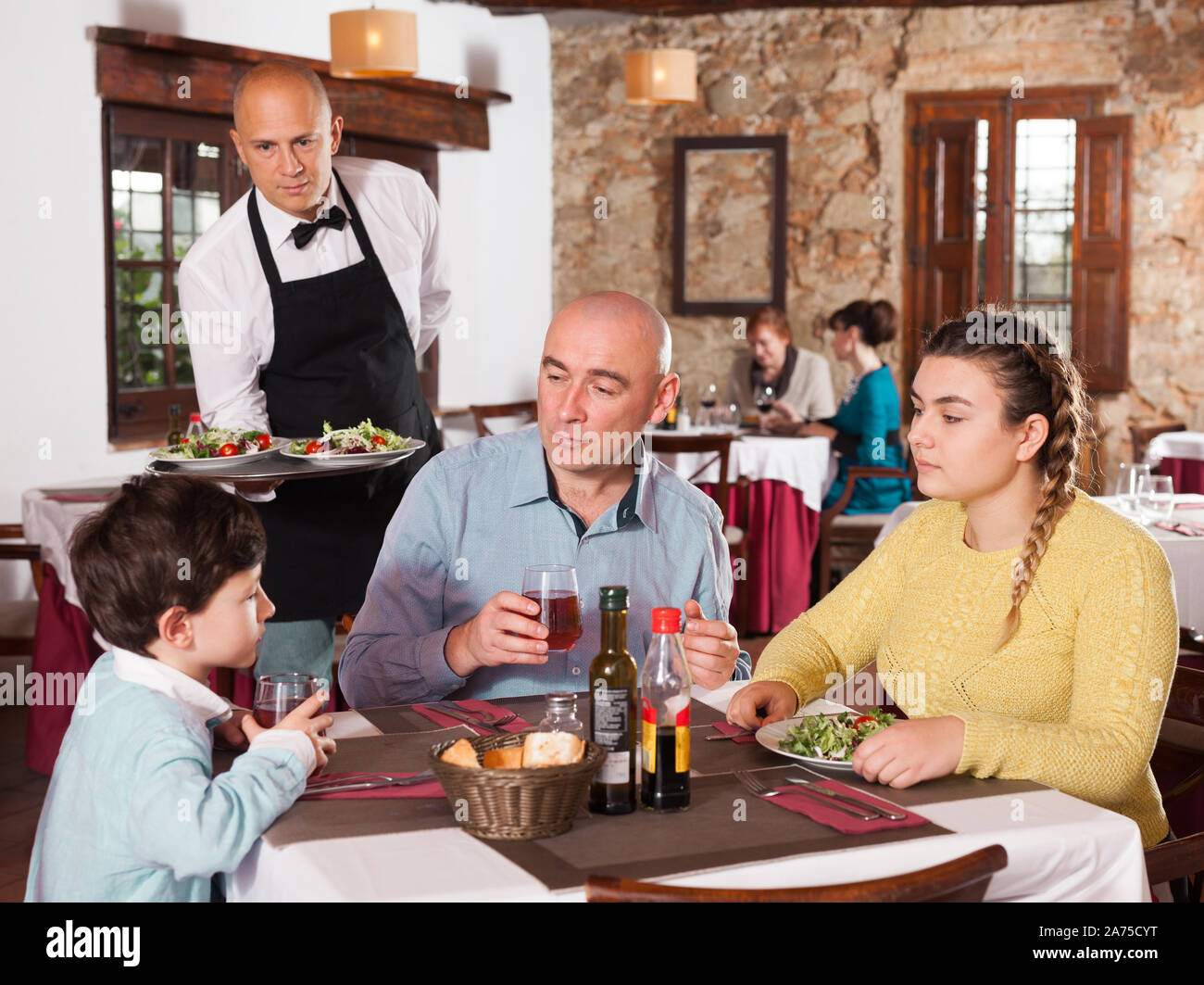 Male waiter holding tray and serving plates of salad to family with ...