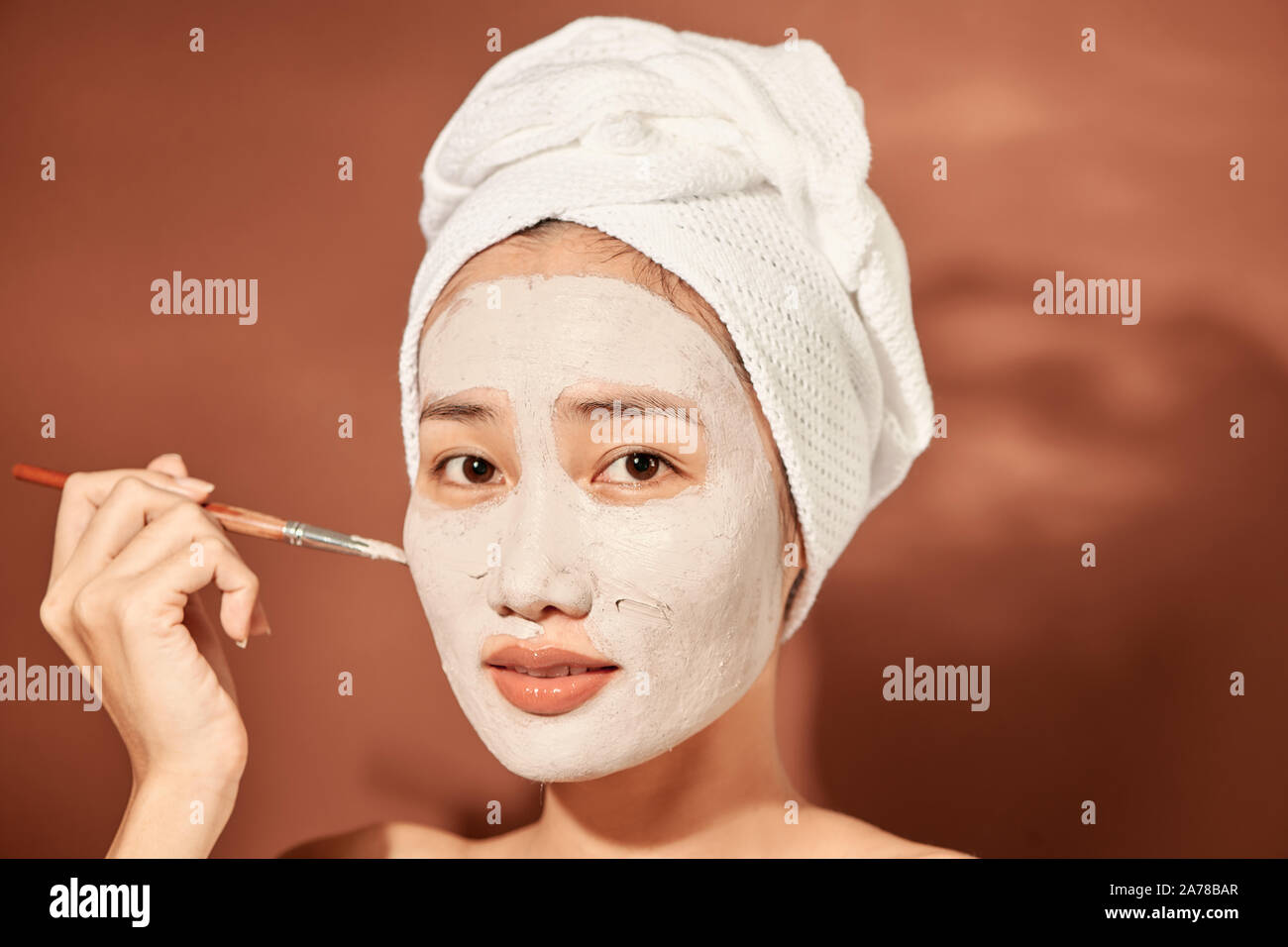 happy woman with a towel on her head apply a cleansing mask on her face ...