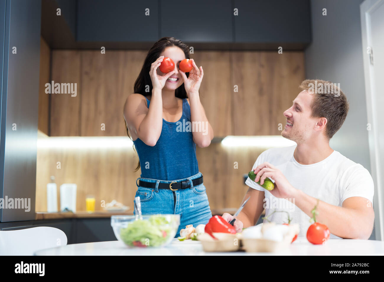 young funny couple cooking breakfast in the kitchen wife holds tomatoes ...