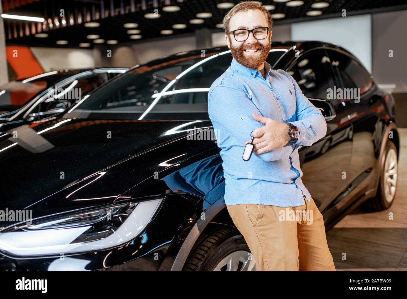 Portrait of a happy young man standing near a new electric car, feeling ...