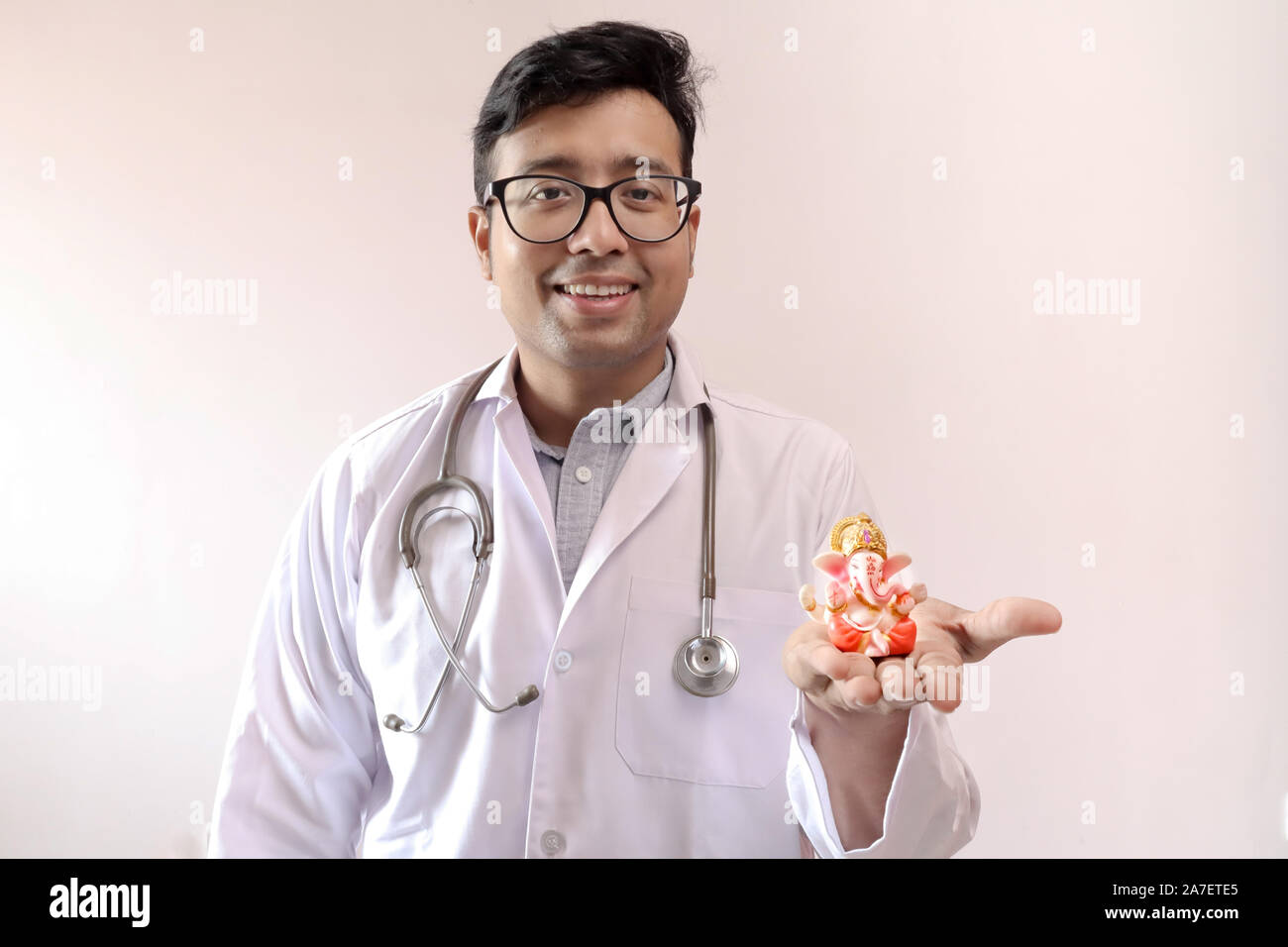 male indian doctor in white coat and stethoscope with a statue of lord ...