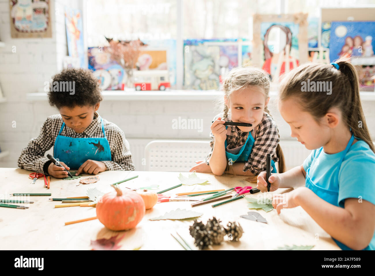 Cute schoolgirl looking at one of classmates drawing face on dry leaf ...