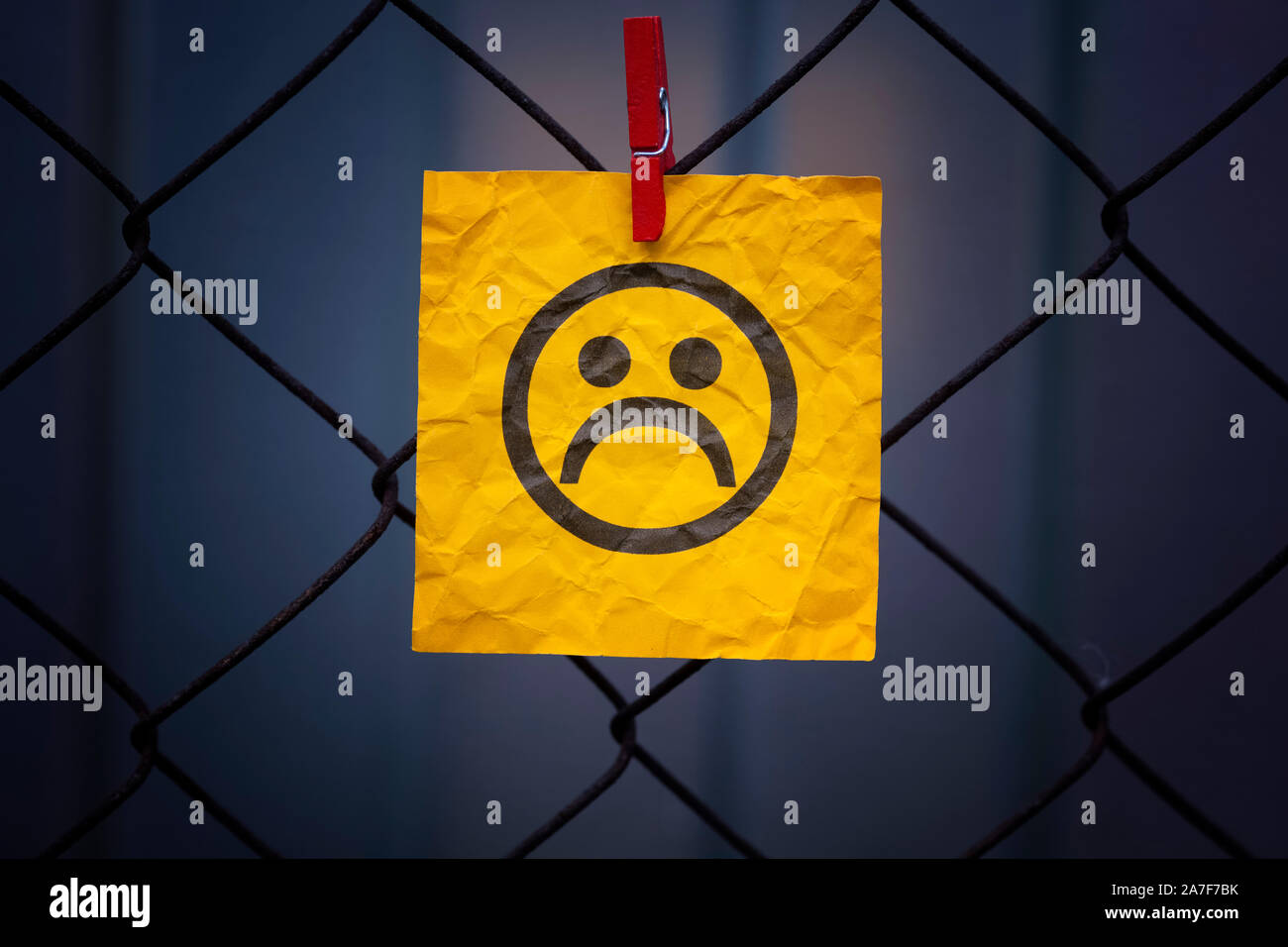 Yellow paper note with sad face hanging on a metal mesh fence. Close up ...