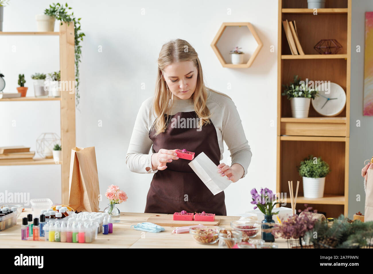 Young woman in apron putting one of handmade soap bars into paper ...