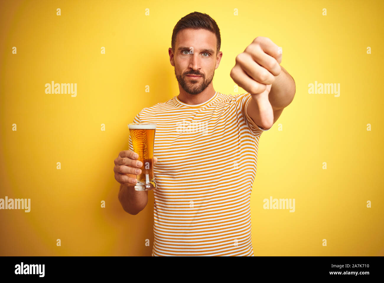 Young handsome man drinking a pint glass of beer over isolated yellow ...