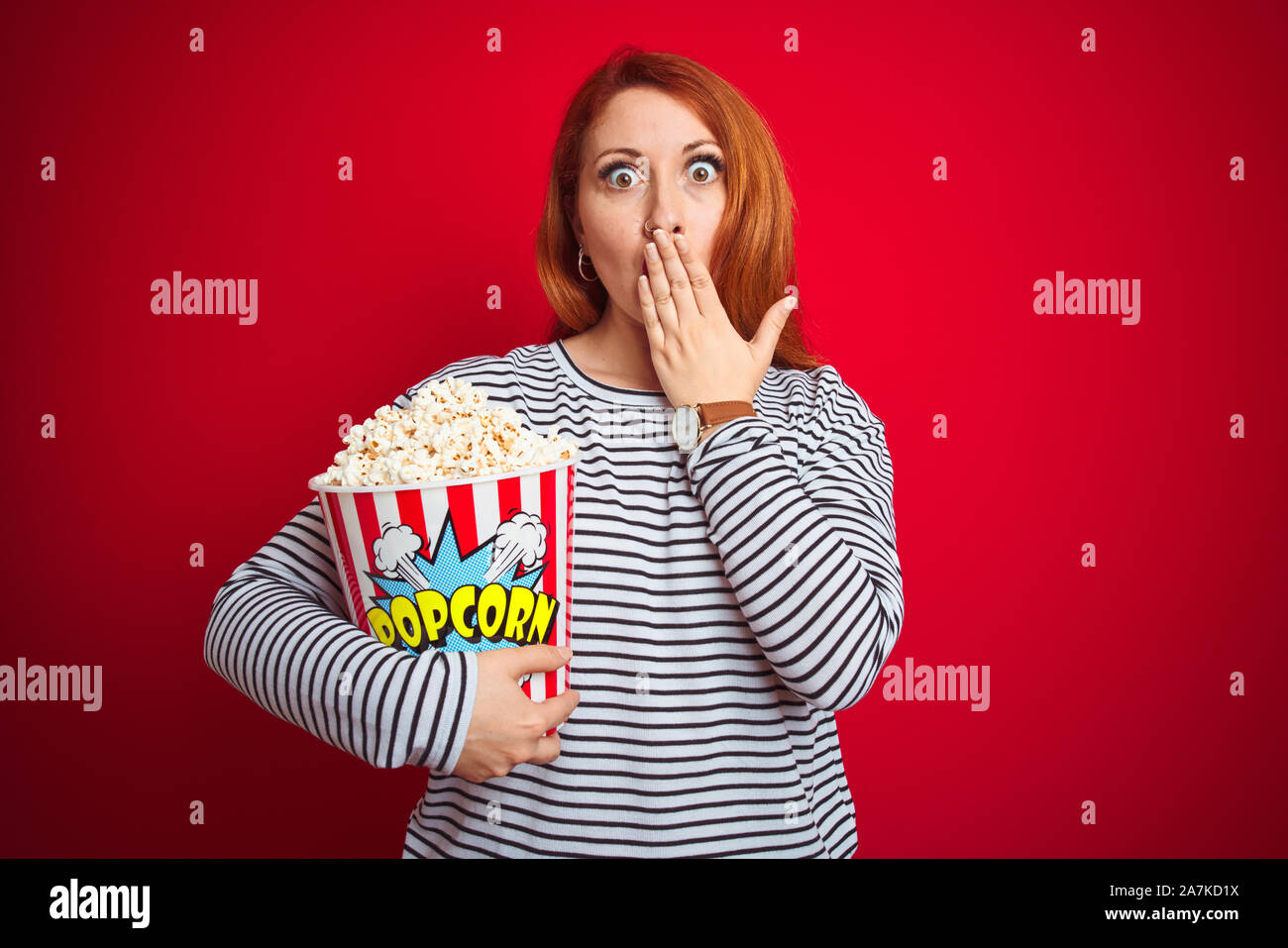 Young beautiful redhead woman eating popcorn over red isolated ...