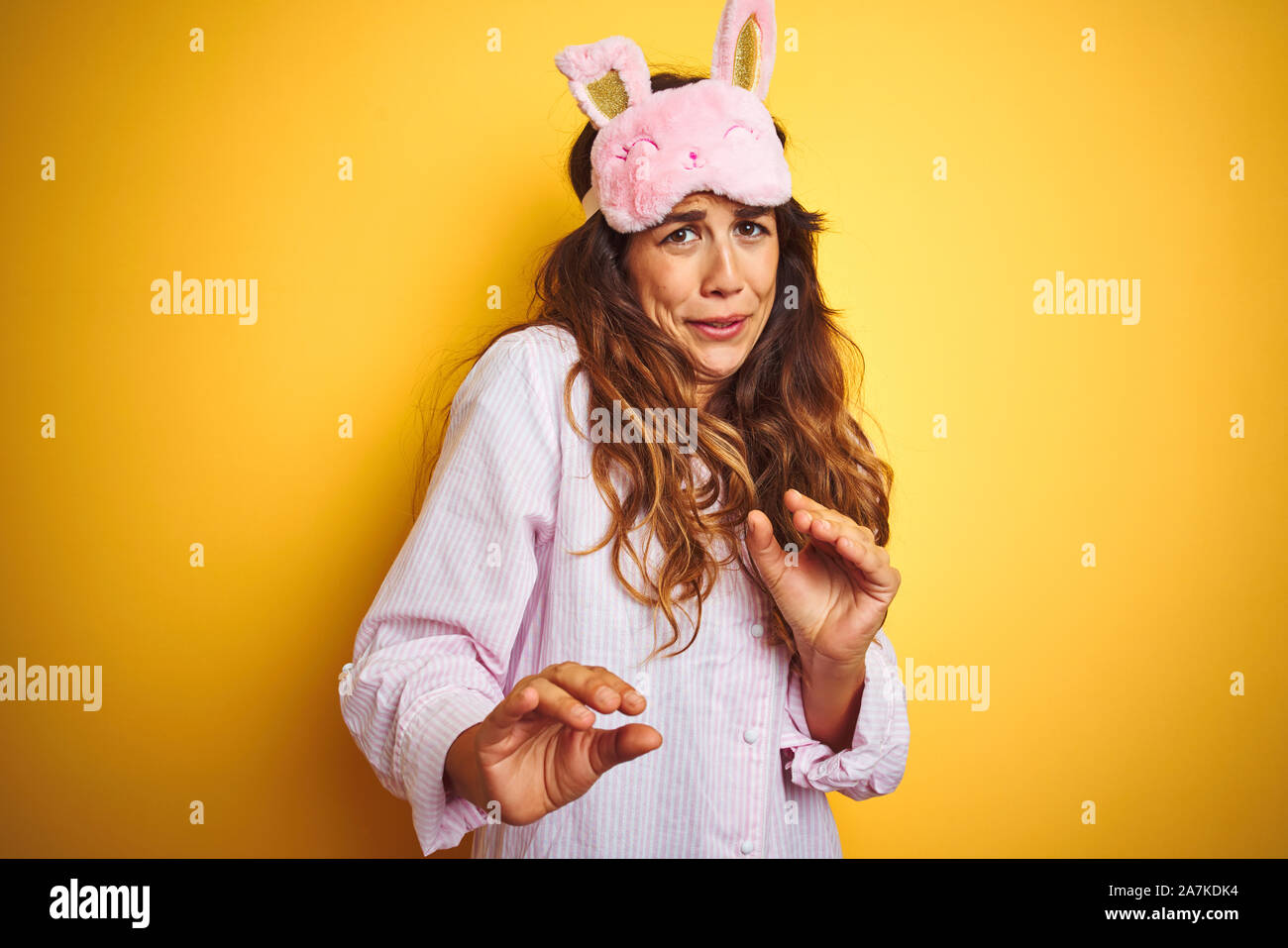 Young woman wearing pajama and sleep mask standing over yellow isolated ...