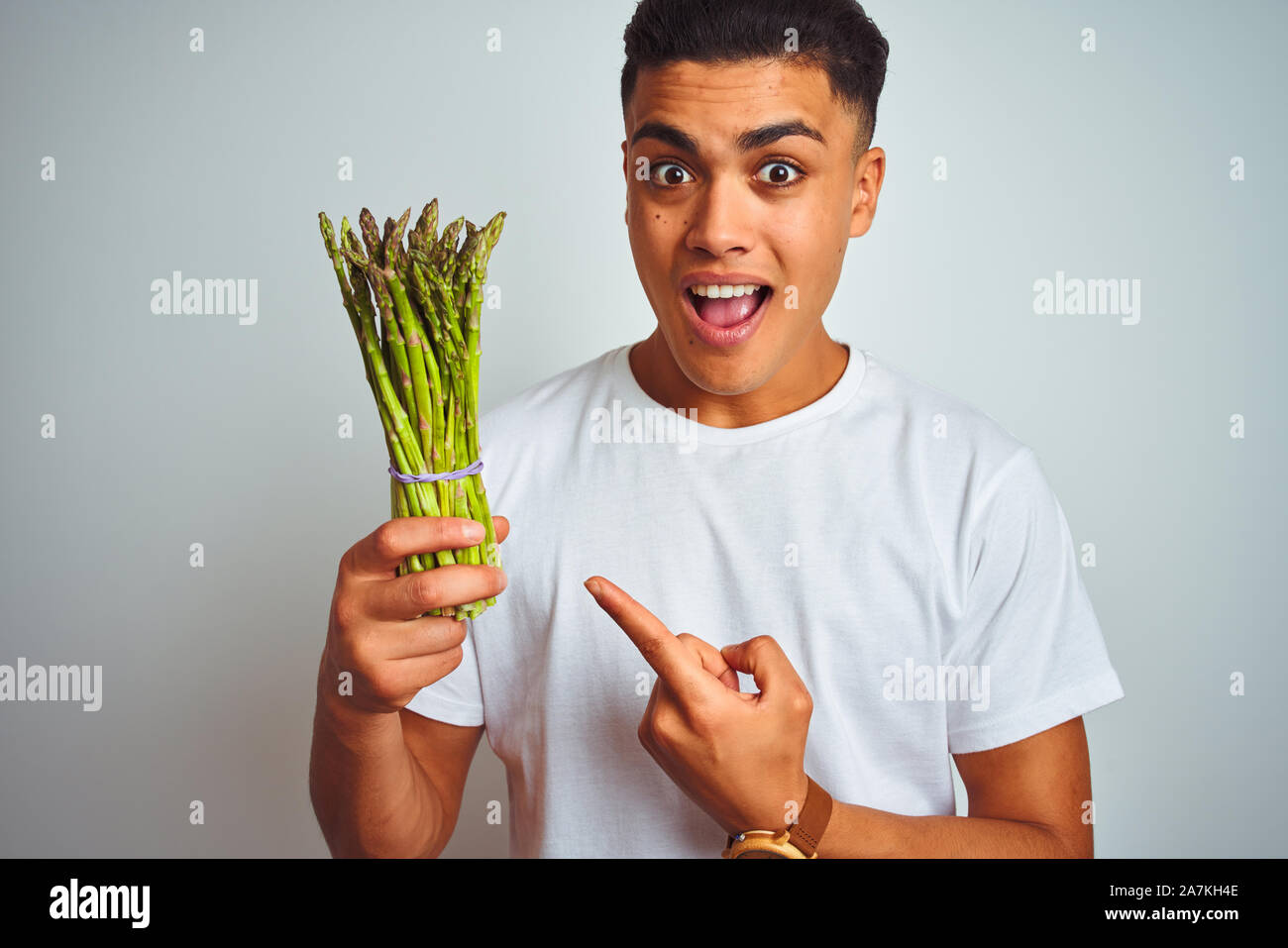 Young brazilian man eating asparagus standing over isolated grey ...