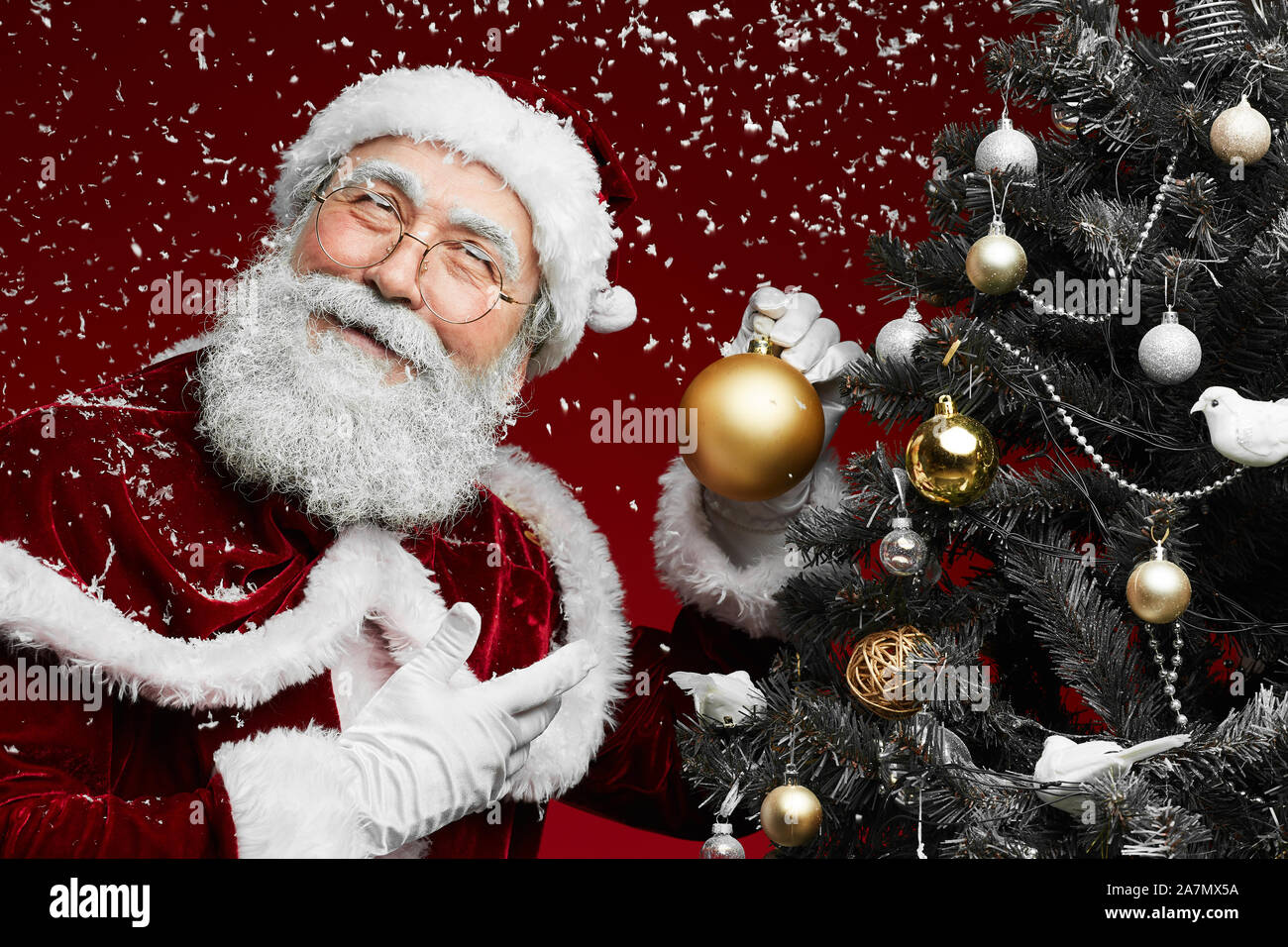 Portrait of cheerful Santa holding golden ornament by Christmas tree ...