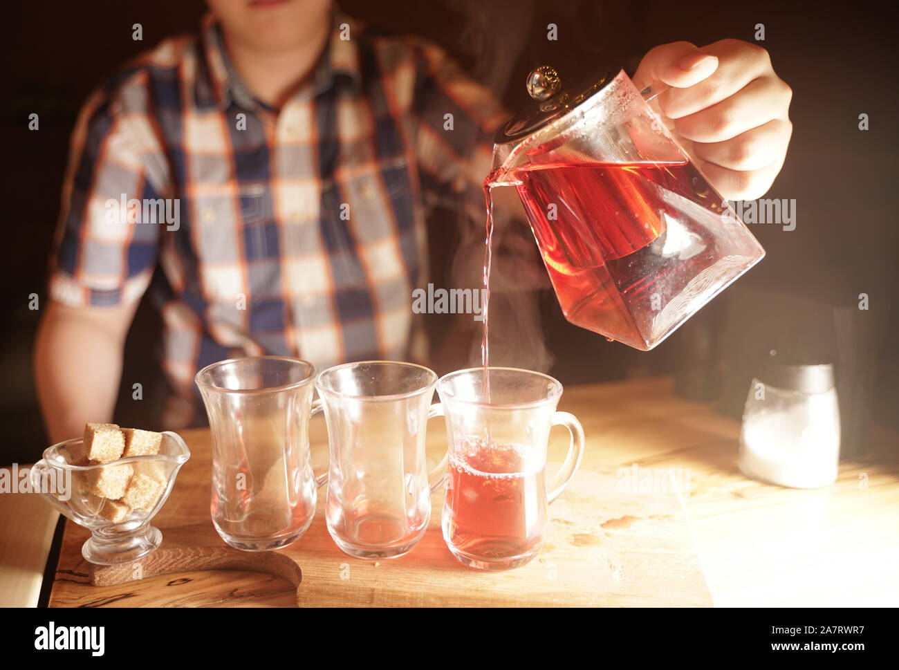 teen boy pours hot tea in transparent teapot into cups on table on dark ...