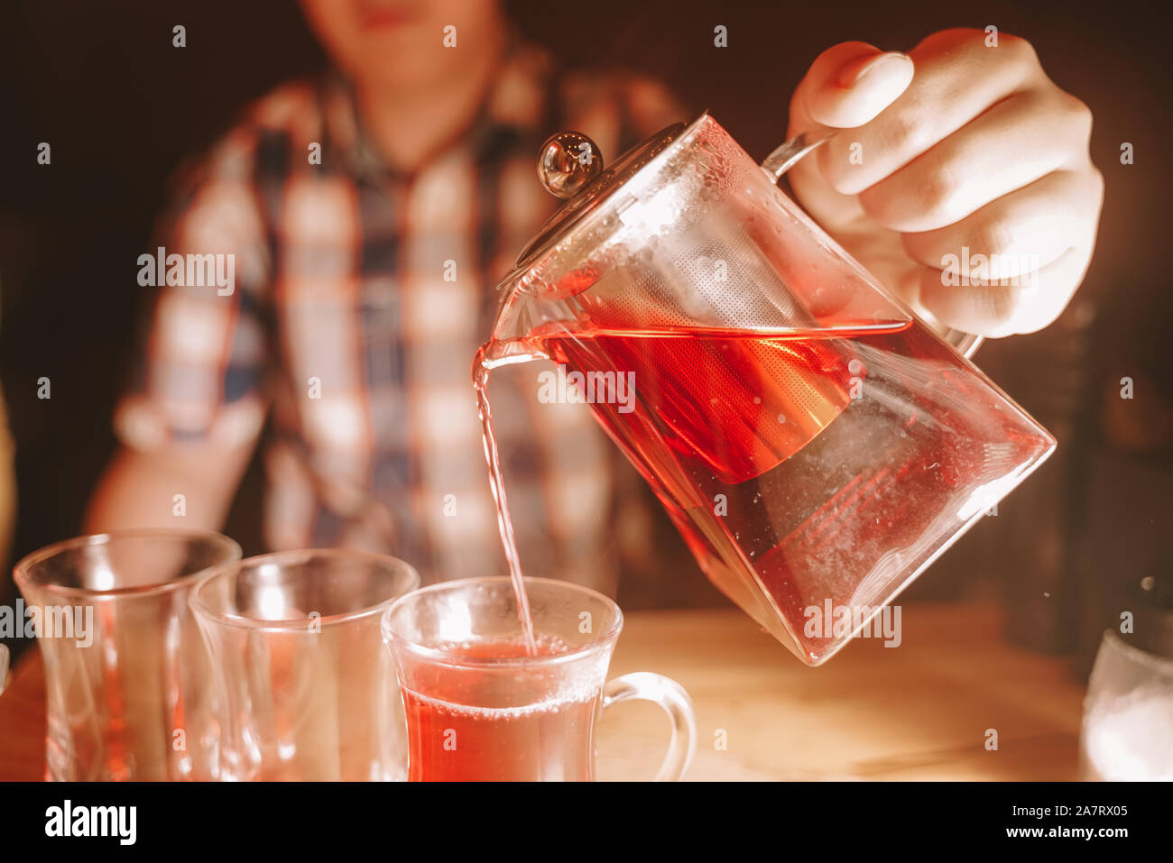 teen boy pours hot tea in transparent teapot into cups on table on dark ...