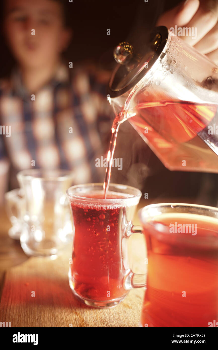 teen boy pours hot tea in transparent teapot into cups on table on dark ...