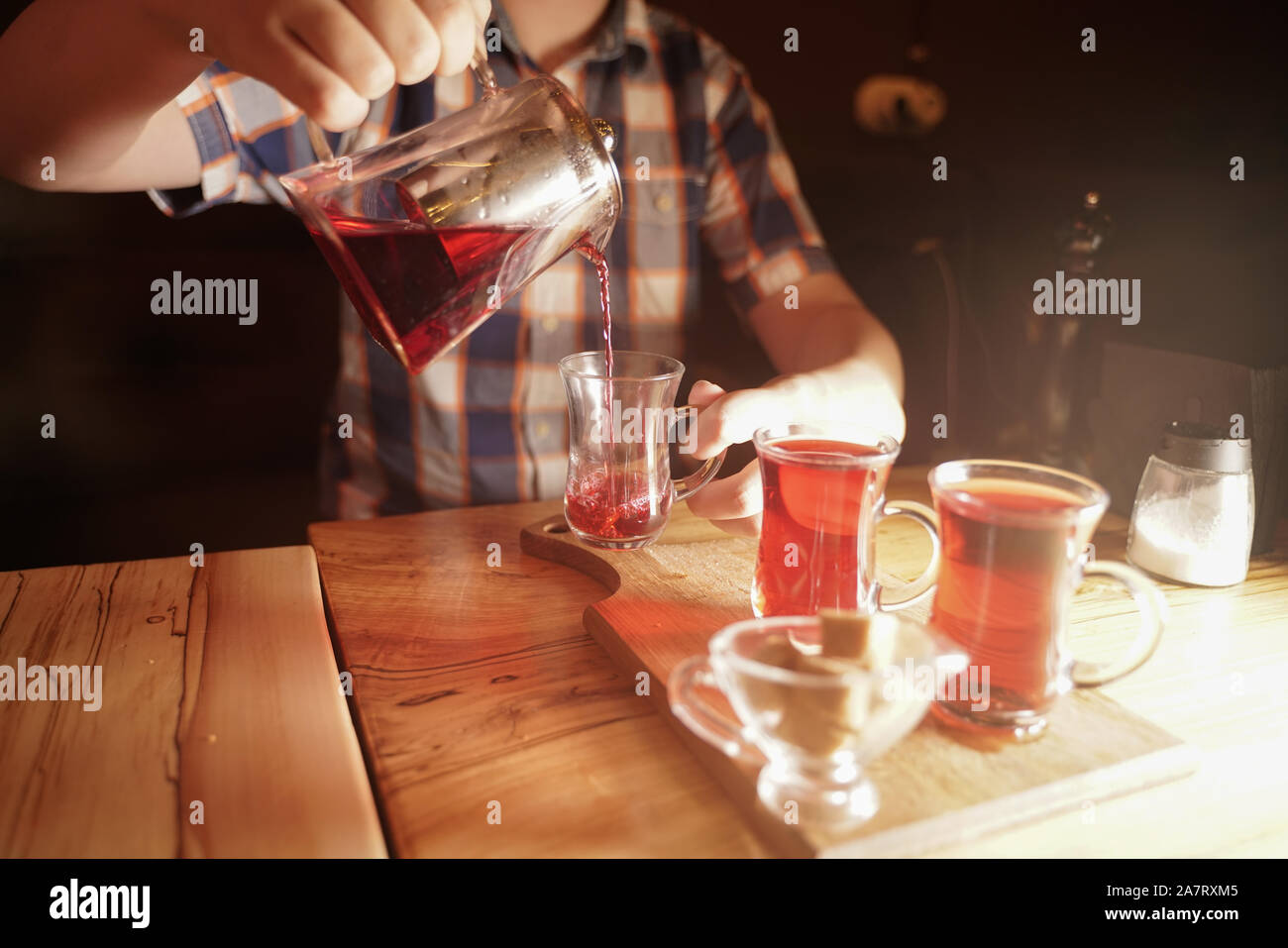 teen boy pours hot tea in transparent teapot into cups on table on dark ...