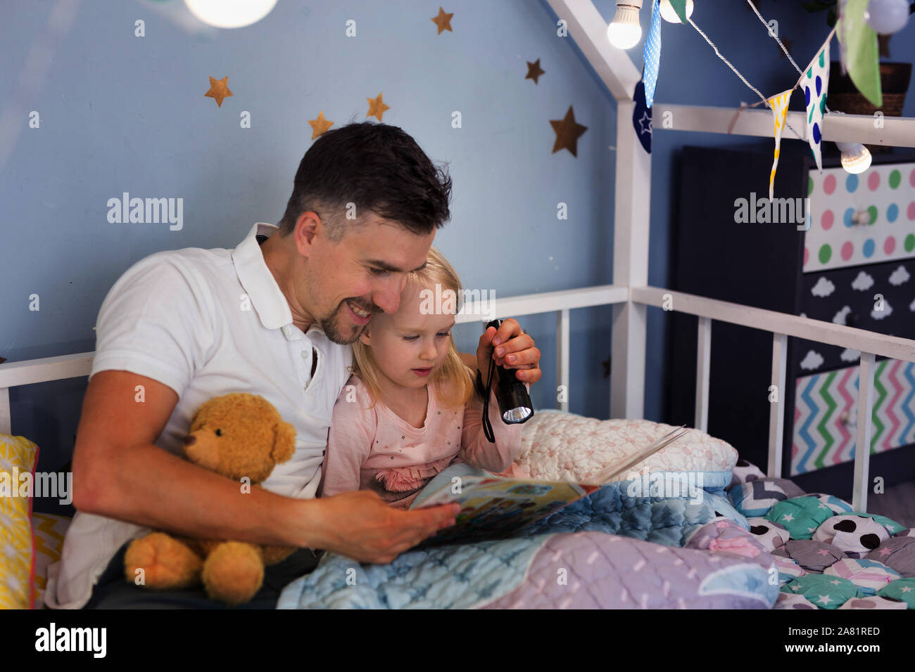 Young handsome father and her daughter reading a book with a flashlight ...