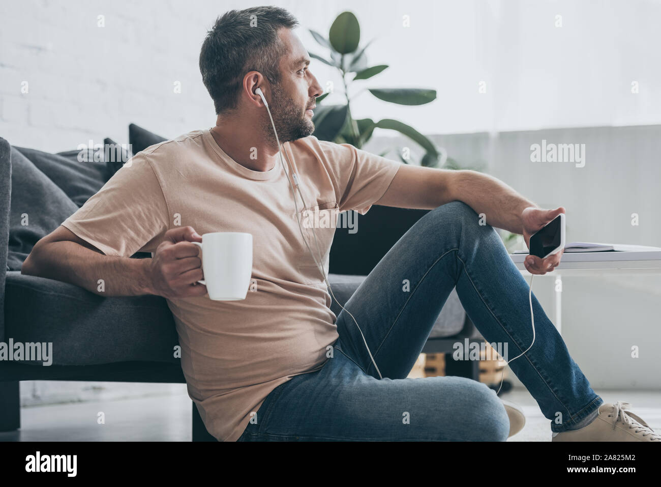 dreamy man looking away while sitting on floor, listening music in earphones and holding coffee ...