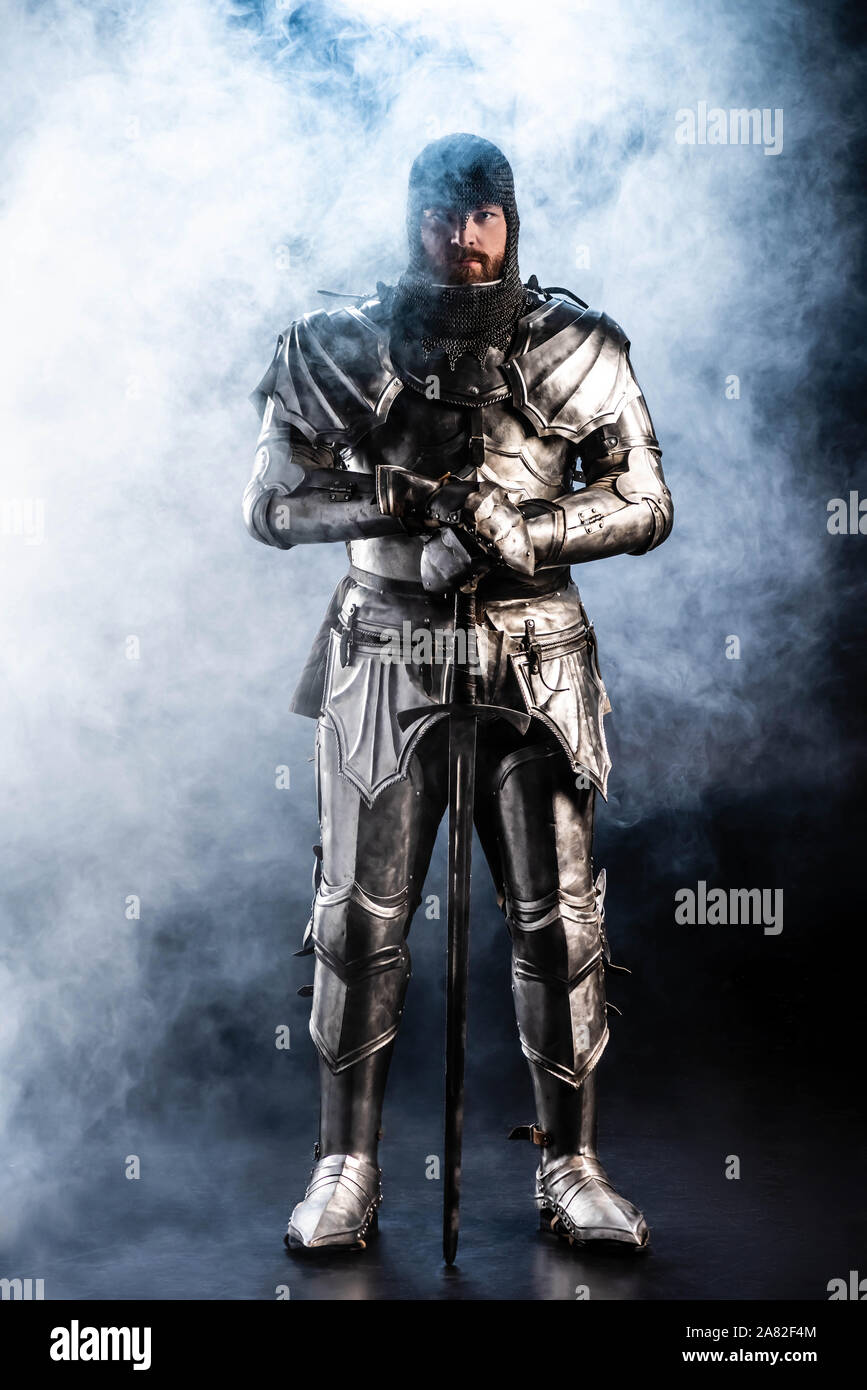 handsome knight in armor looking at camera and holding sword on black ...