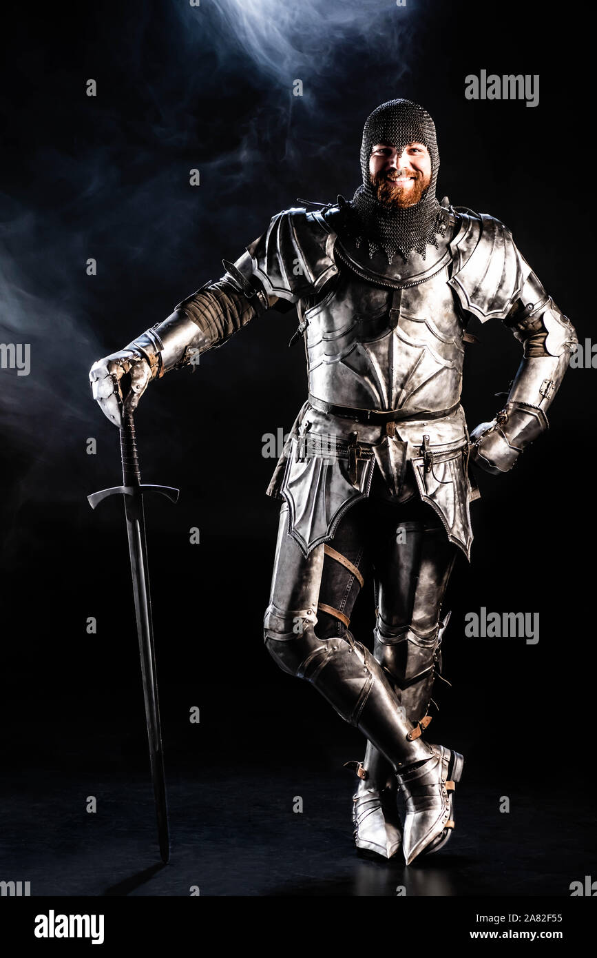 smiling knight in armor looking at camera and holding sword on black ...
