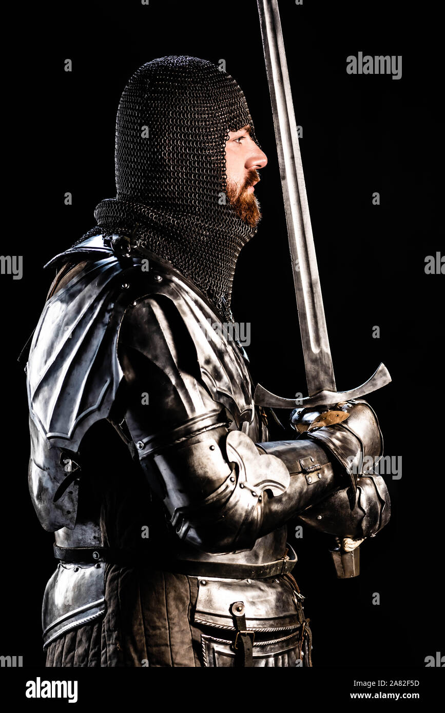 side view of handsome knight in armor holding sword isolated on black ...