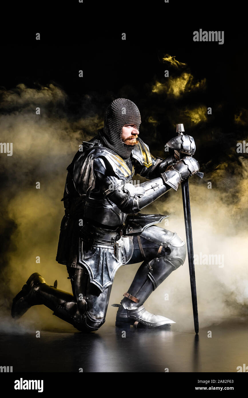 handsome knight in armor holding sword and bend knee on black ...
