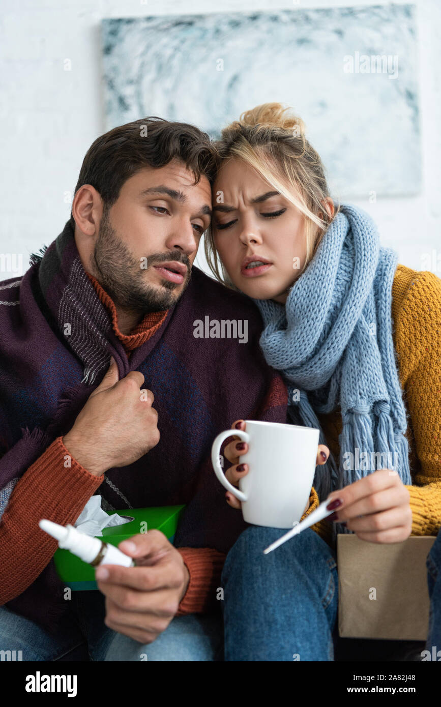 sick couple with fever holding thermometer, hot drink and nasal spray ...